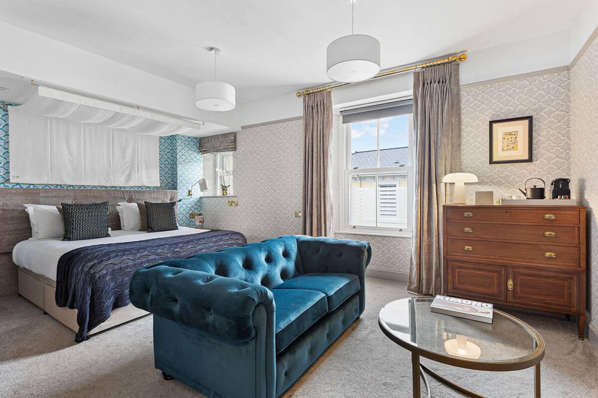Quarto com uma cama, um sofá azul, cômoda de madeira e mesa de vidro.