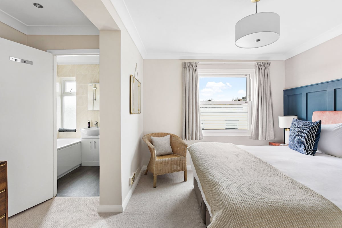 Quarto com cama, uma cadeira, janela com cortinas, mesa de cabeceira com abajur e vista para o exterior.