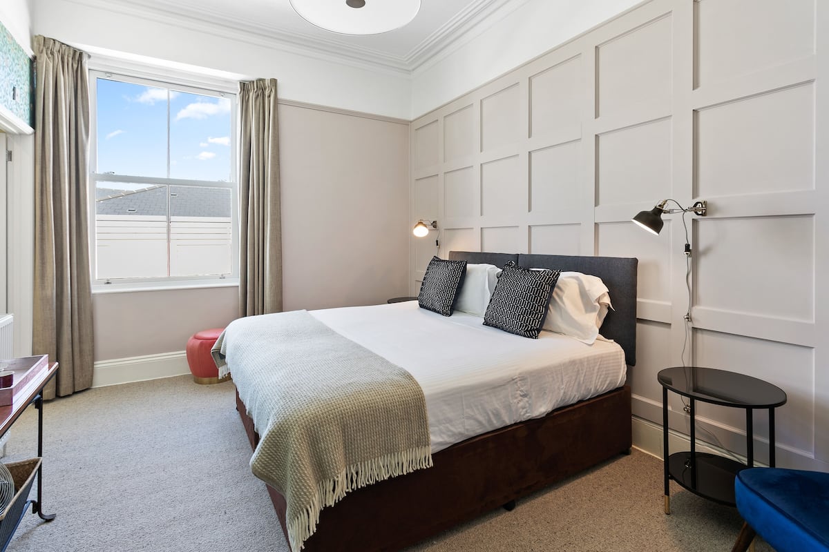 Quarto com uma cama grande, uma mesa de cabeceira, uma luminária e vista para o exterior através de uma janela.