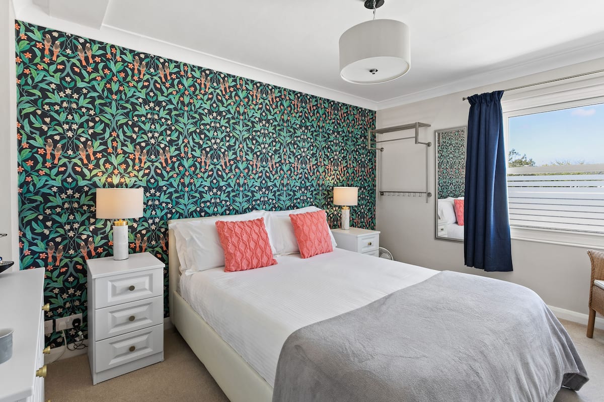 Quarto com papel de parede floral, cama com roupa de cama branca e duas almofadas rosa, mesas de cabeceira com abajures, janela com cortinas e vista para o exterior.