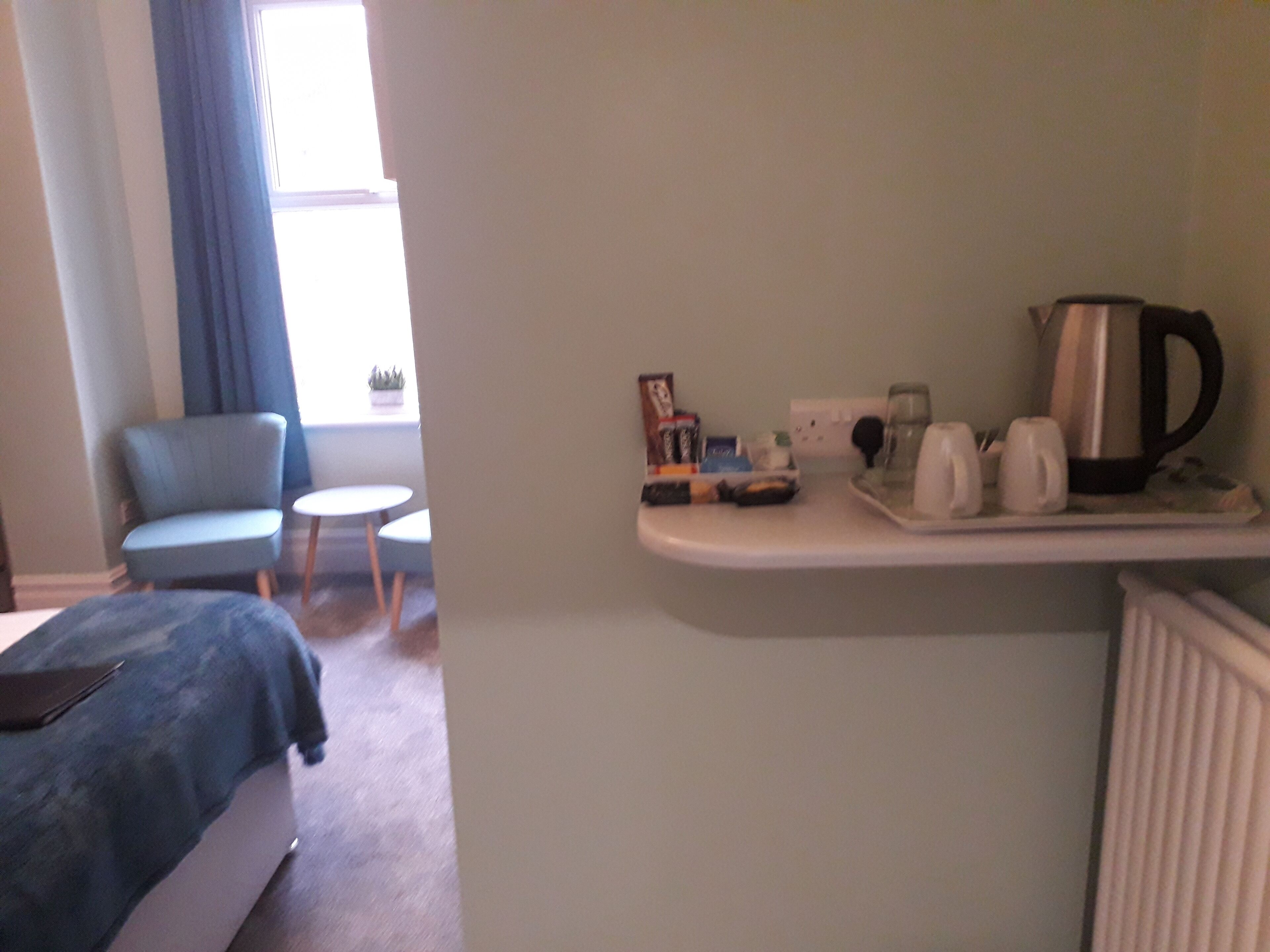 Quarto de hotel com cama, poltrona, mesa pequena, cozinha compacta com chaleira, xícaras e bandeja.