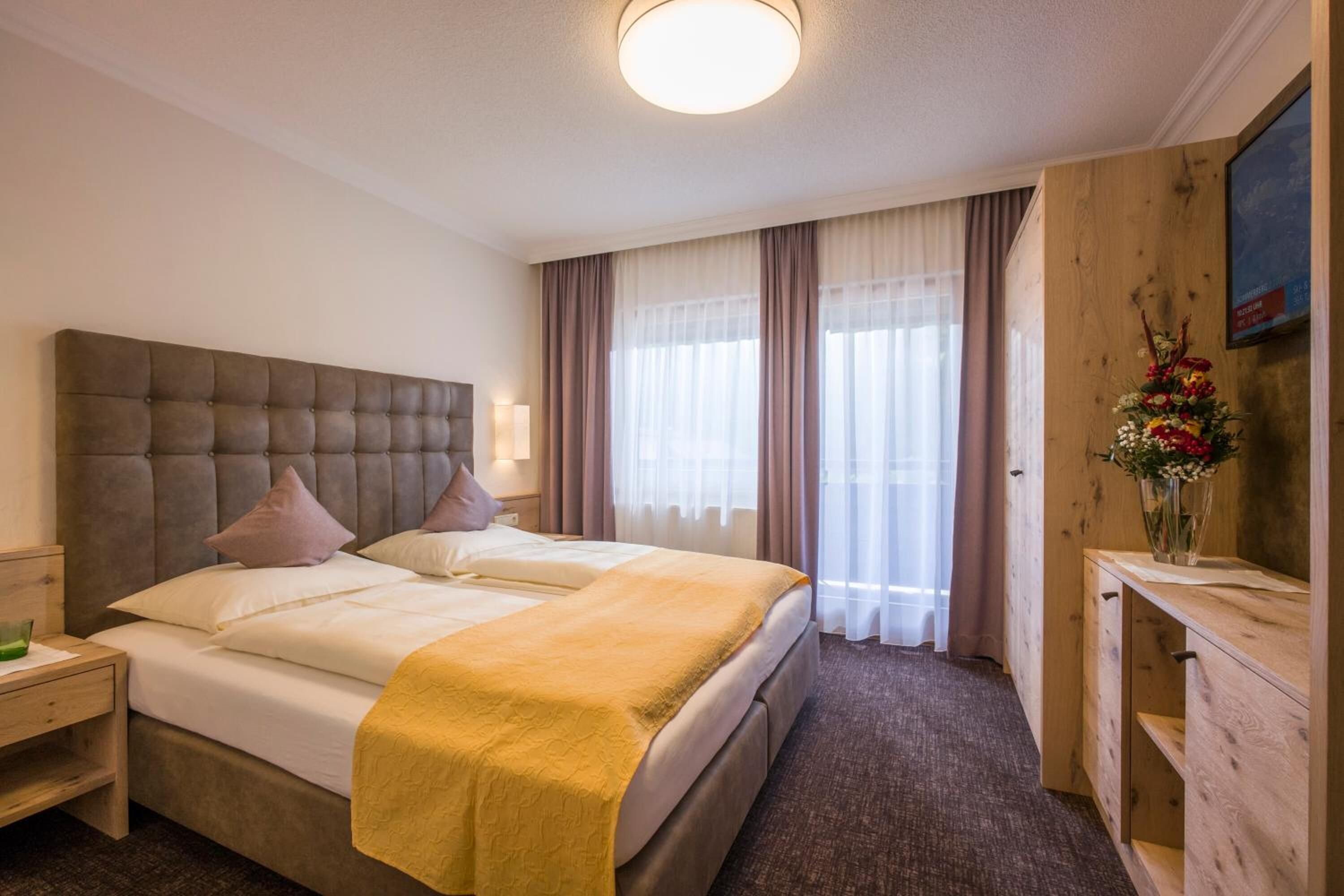 Quarto de hotel com uma cama grande, cabeceira de madeira, mesa de cabeceira, cómoda de madeira, espelho e janela com cortinas.