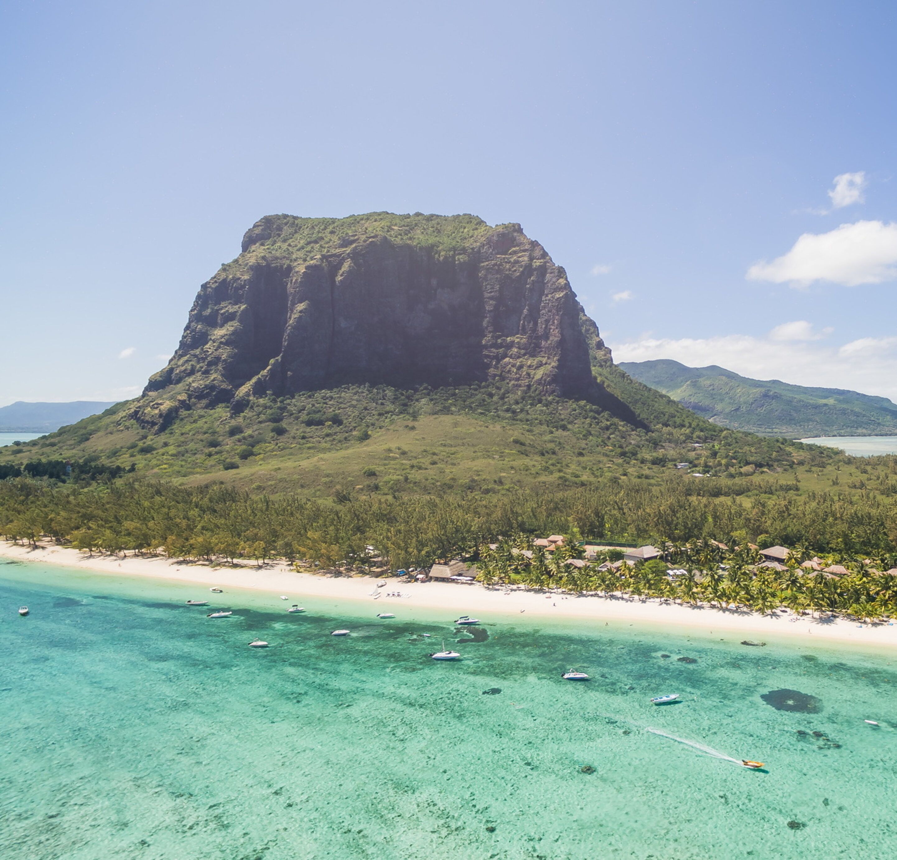Paisagem costeira com uma montanha imponente, águas turquesa cristalinas e uma praia com edifícios e palmeiras.