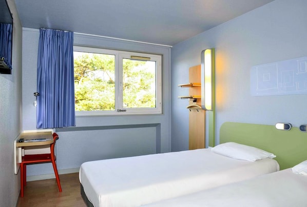 Quarto de hotel com duas camas, uma escrivaninha, uma cadeira, uma janela com cortinas e um candeeiro fixado na parede.
