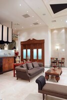 Uma sala de estar espaçosa com sofá, mesa de centro e mesa de jantar com cadeiras. Há uma mesa de recepção com uma placa que diz "Coffee Shop". O ambiente possui pé direito alto com iluminação embutida e uma grande janela.