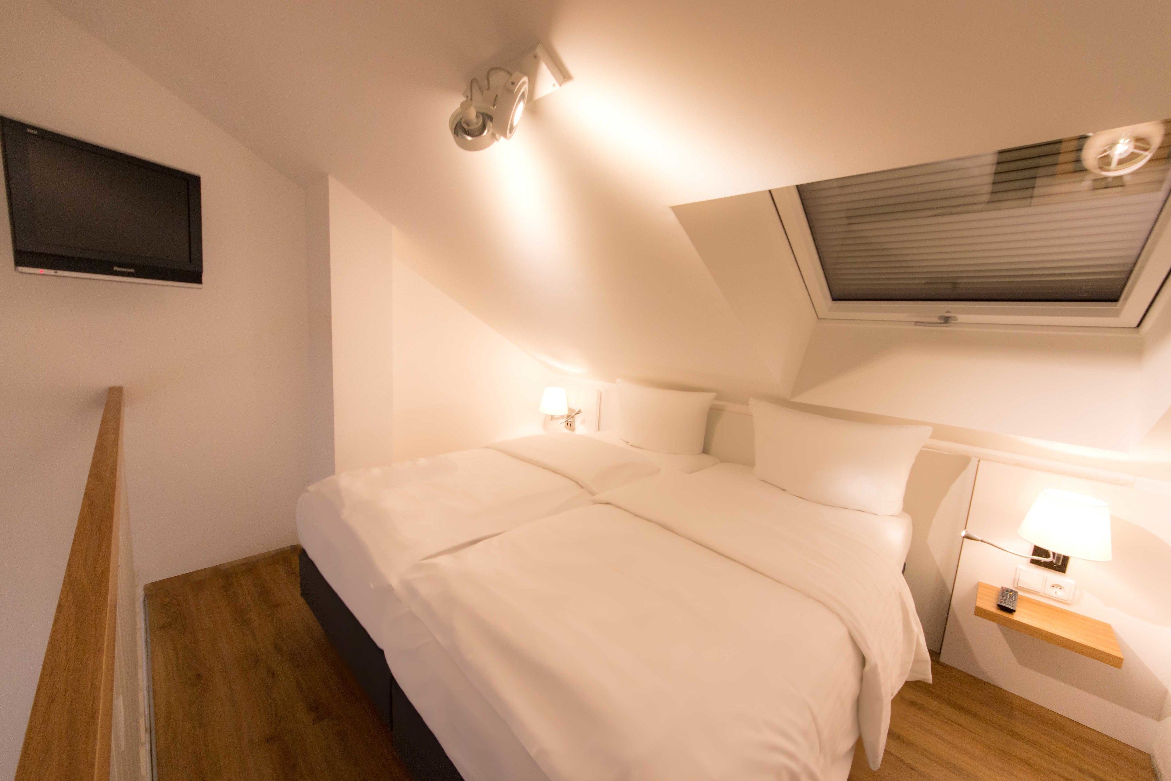 Quarto com cama, mesa de cabeceira com abajur, TV fixada na parede e janela com persianas.