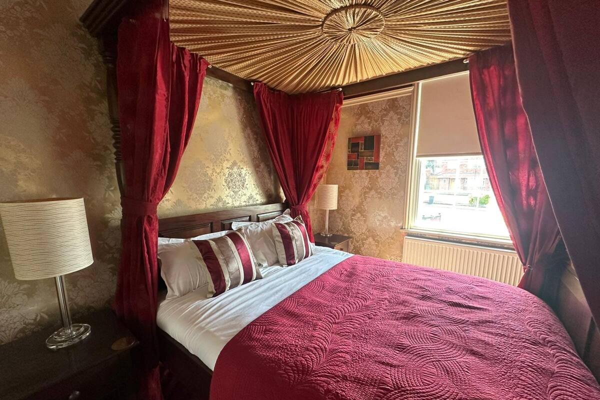Quarto com cama, cortinas vermelhas, luminária e janela com vista.