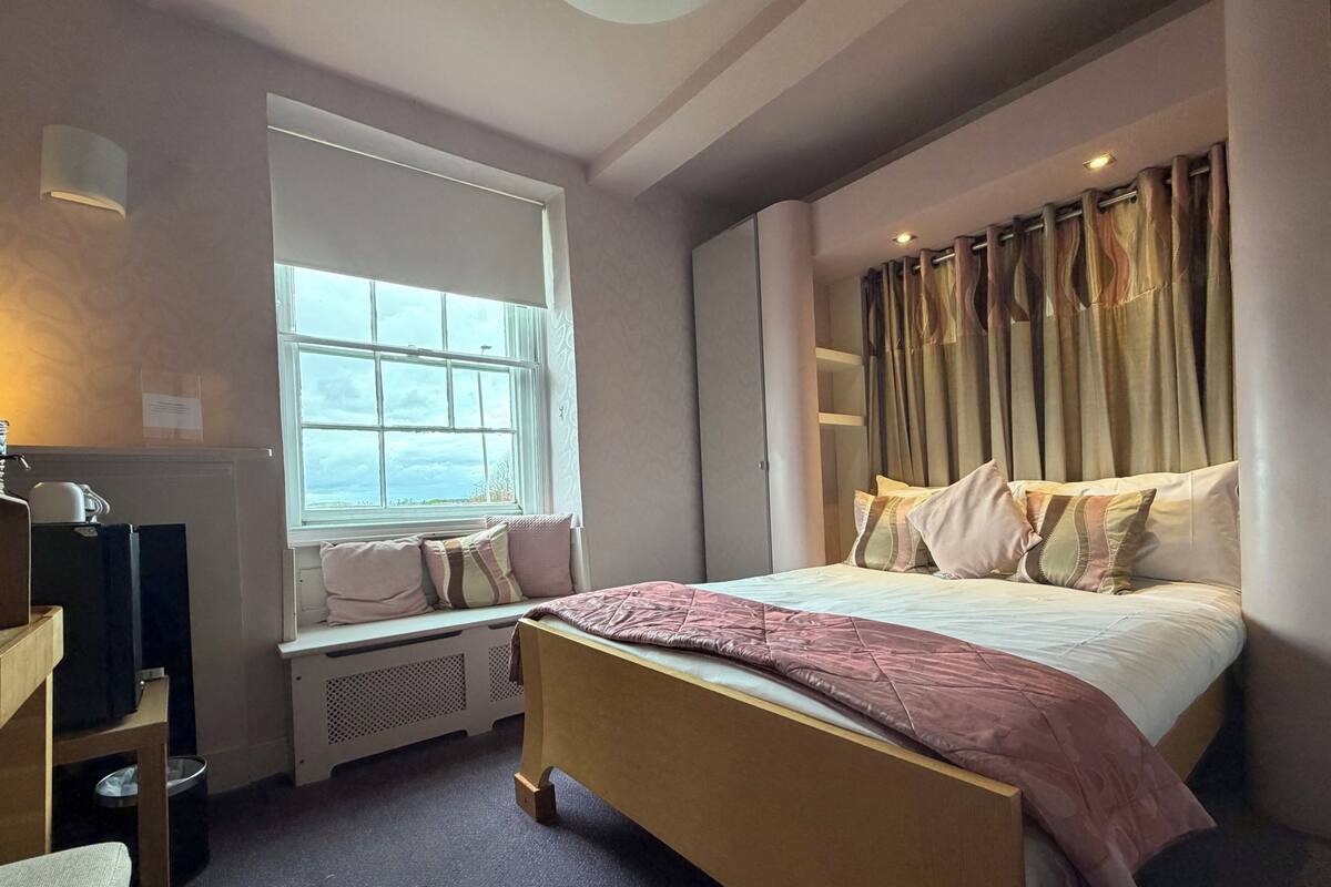 Um quarto com uma cama, uma janela com persianas, um radiador e uma escrivaninha com luminária.