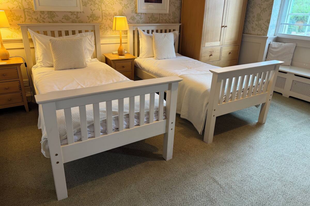 Quarto com duas camas de solteiro, uma mesa de cabeceira com abajur, guarda-roupa de madeira e papel de parede floral.