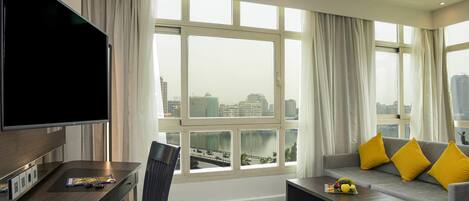Quarto de hotel com uma janela grande, televisão de tela plana, mesa de jantar, sofá com almofadas amarelas e mesa de centro com frutas.
