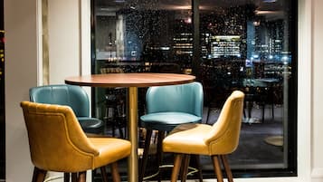 Um interior moderno com uma mesa redonda e quatro cadeiras, uma janela ampla com vista para a cidade, e uma área de bar visível através da janela.