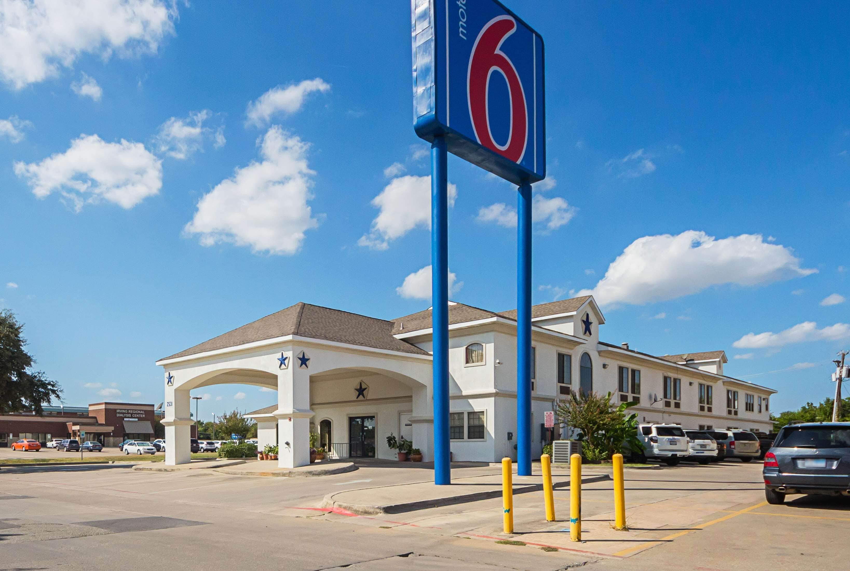 Um motel com uma grande placa azul indicando a suíte número 6, uma entrada coberta e vários carros estacionados na frente.