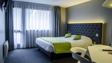 Quarto de hotel com cama, escrivaninha, cadeira, janela com cortinas e luminária na parede.