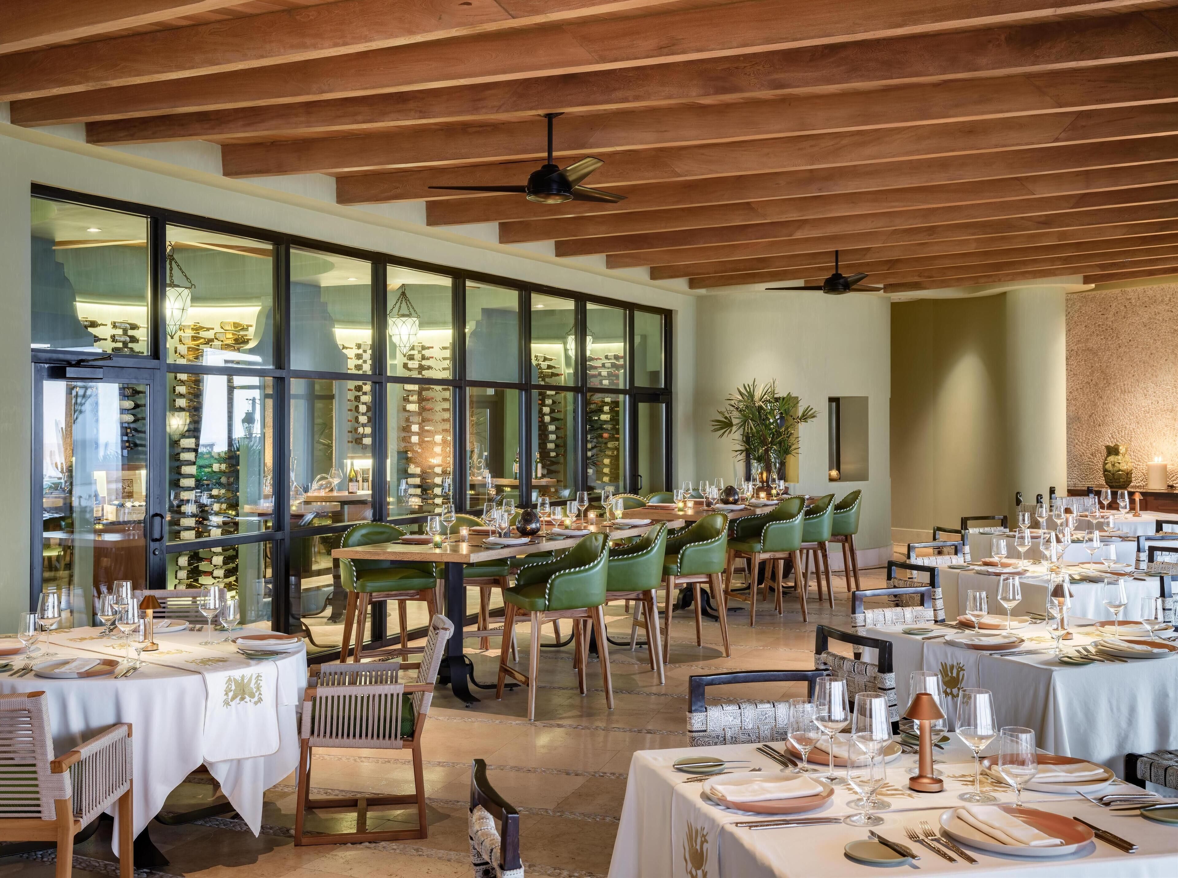 Um restaurante com várias mesas dispostas para refeições, apresentando um interior moderno com vigas de madeira no teto e grandes janelas.