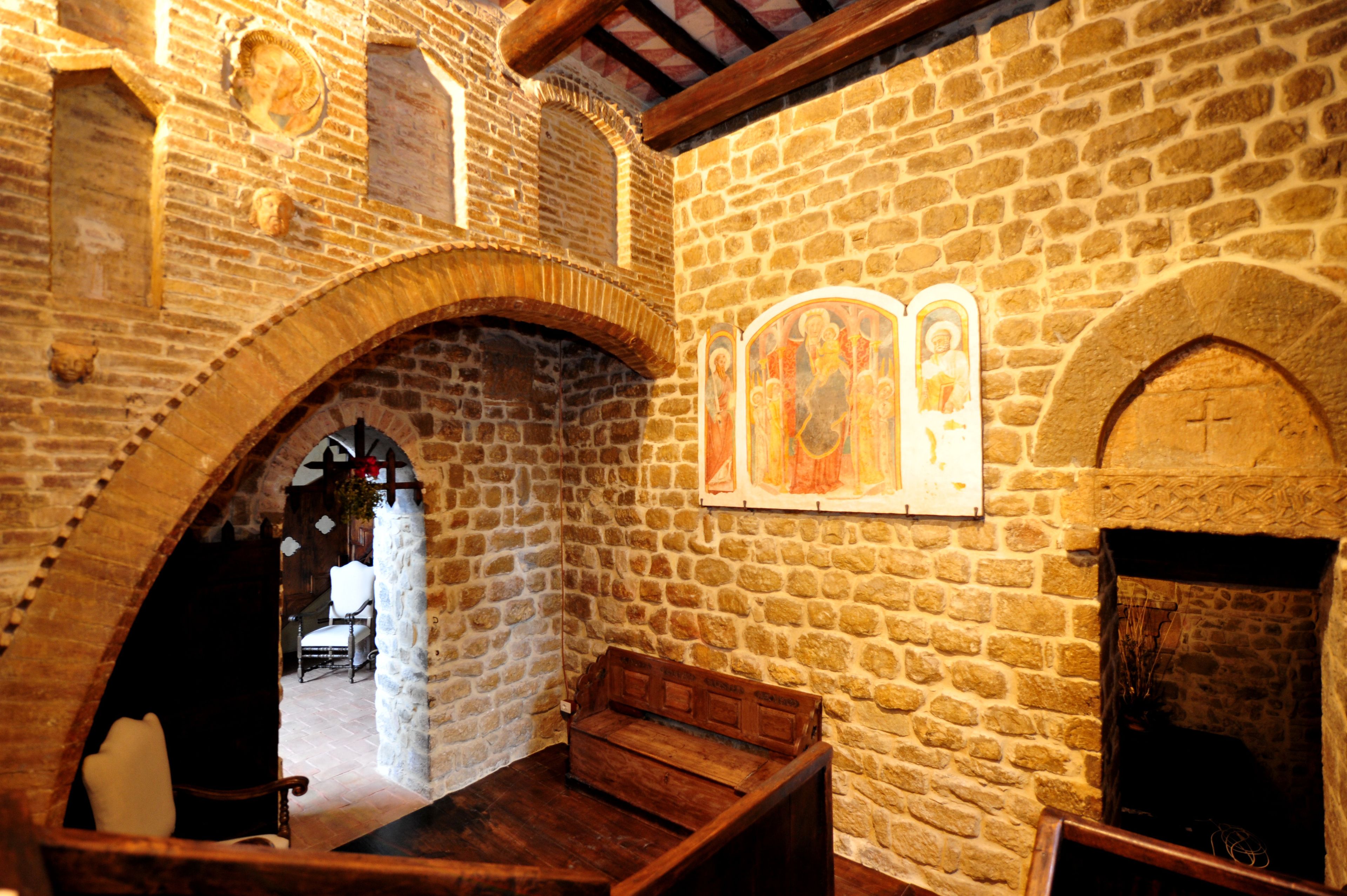 Quarto com paredes de pedra, um banco de madeira, uma lareira e uma porta que dá para outro aposento.