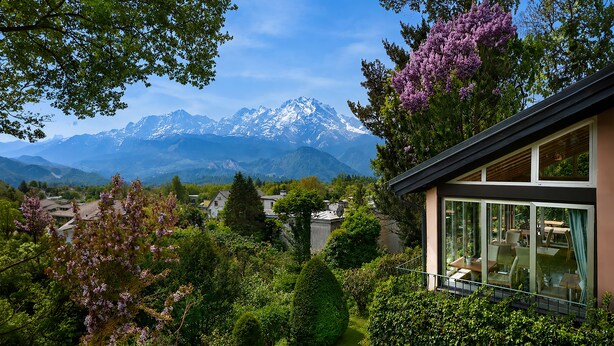 Um edifício moderno com grandes janelas, um jardim bem cuidado e vista para montanhas nevadas.