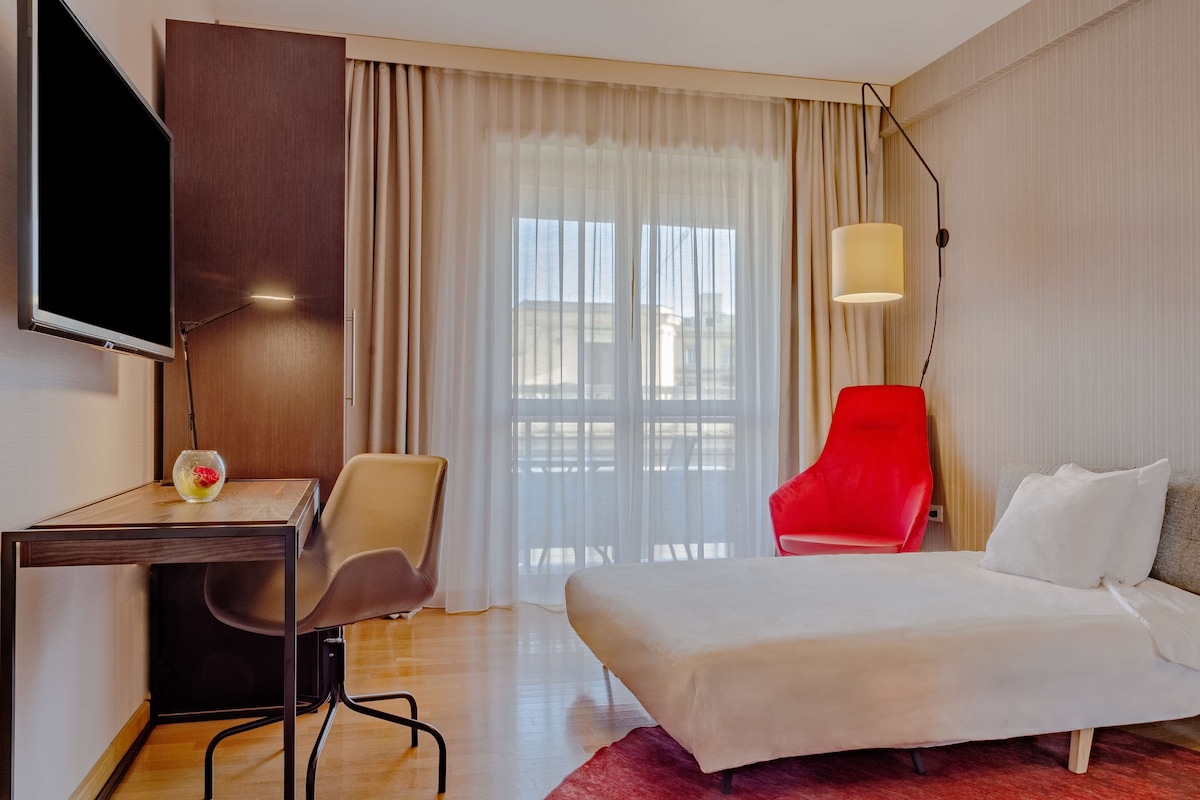 Quarto de hotel com cama, escrivaninha, cadeira, televisão, poltrona vermelha e luminária.