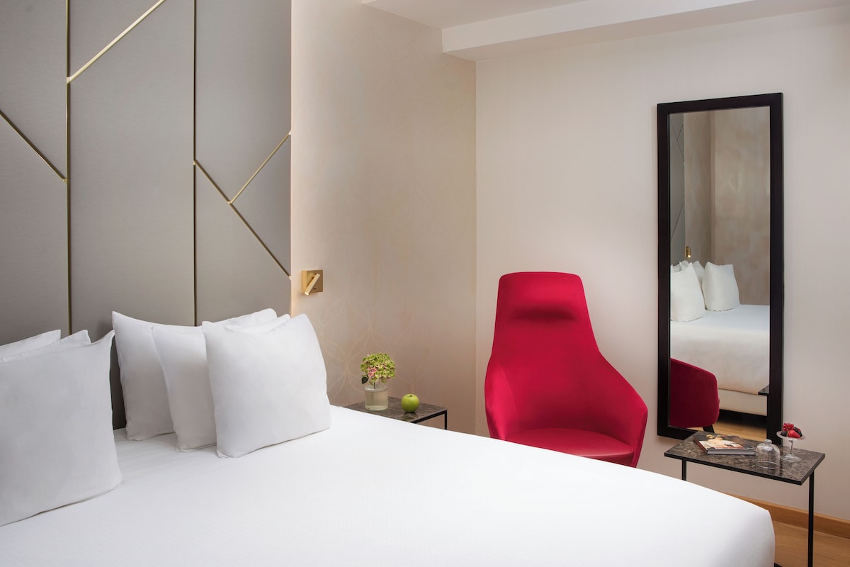 Quarto de hotel com cama, almofadas brancas, uma cadeira vermelha, uma mesinha com um vaso e uma maçã, e um espelho grande refletindo a cama.