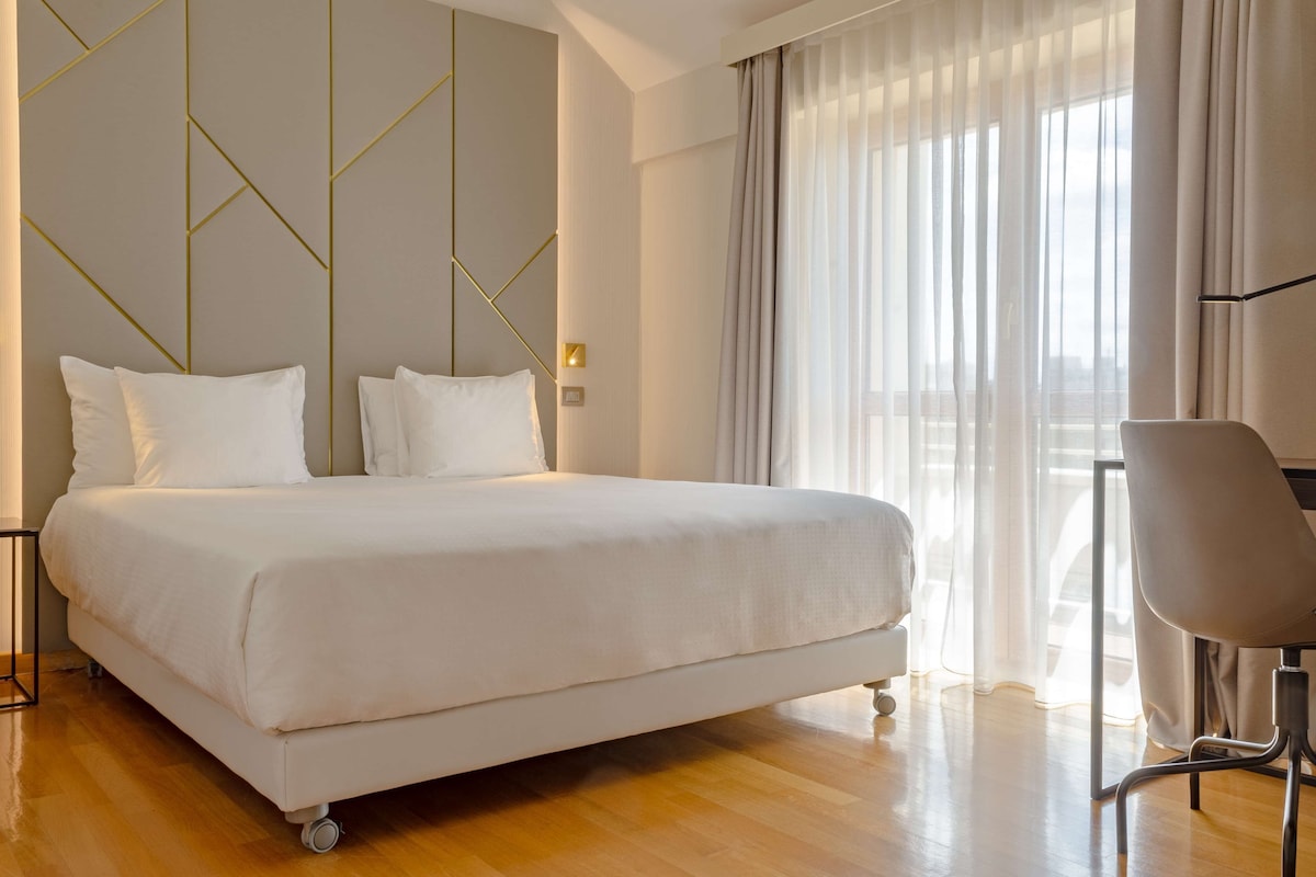 Quarto de hotel moderno com uma cama grande, uma cadeira e vista da cidade através de cortinas leves.