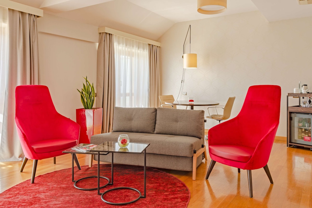 Uma sala de estar moderna com um sofá cinzento, duas poltronas vermelhas, uma mesa de centro de vidro e um tapete vermelho.
