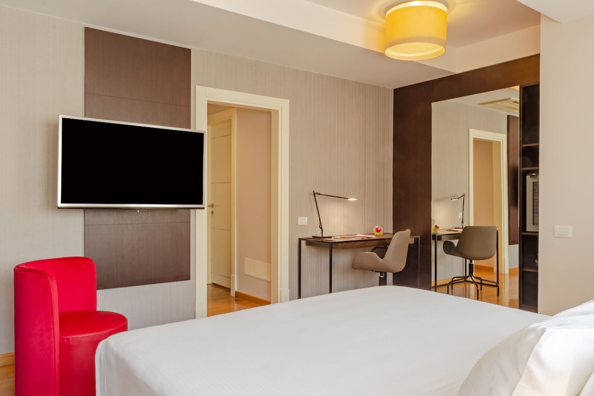 Quarto de hotel com uma cama, televisão presa à parede, secretária com cadeira e uma cadeira vermelha.