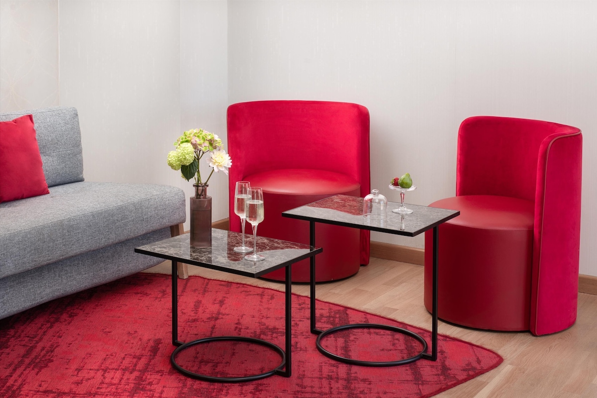 Uma sala de estar moderna com um sofá cinza, cadeiras vermelhas, uma mesa pequena com dois copos e um vaso com flores.