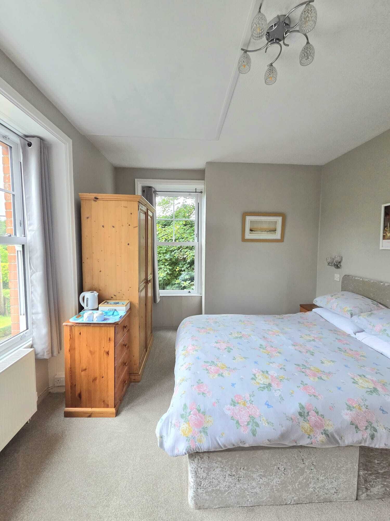 Um quarto com uma cama, um guarda-roupa de madeira, uma pequena mesa com uma chaleira e uma janela com cortinas.