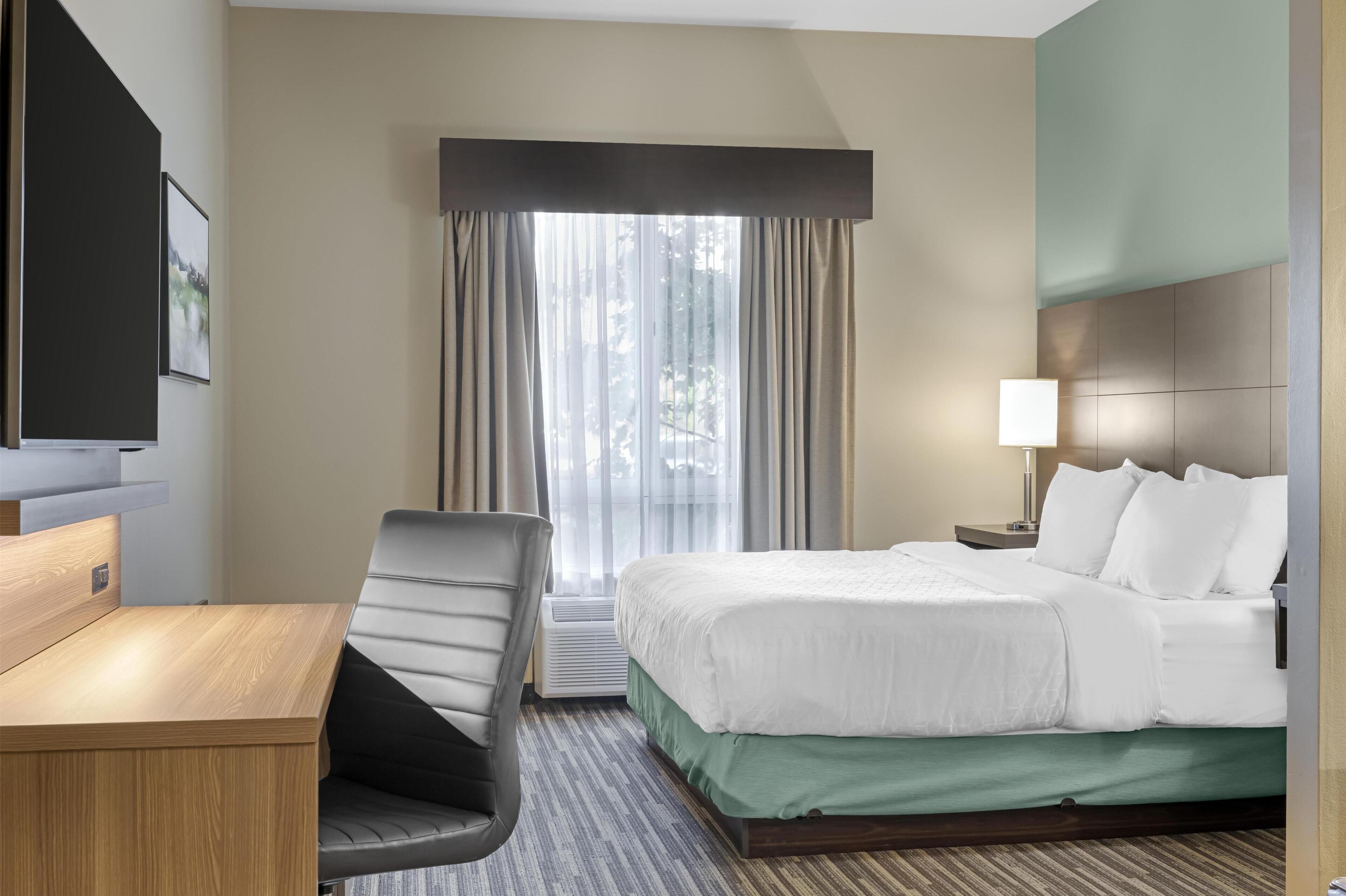 Quarto de hotel com cama, escrivaninha, cadeira, mesa de cabeceira e janela com cortinas.