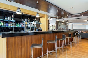 Uma área de bar moderna com balcão de madeira, vários banquetes e variedade de garrafas e copos nas prateleiras.