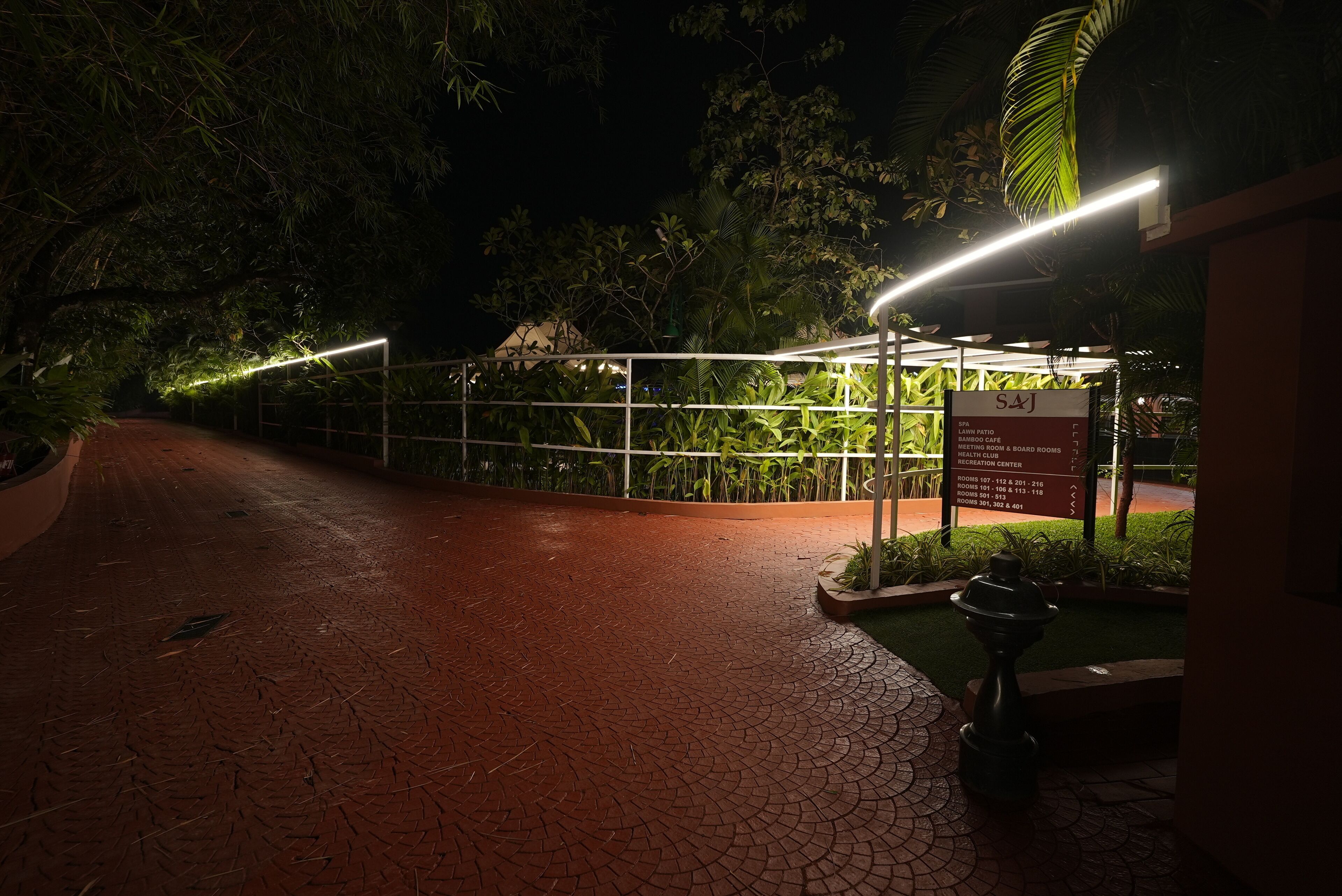 Um caminho pavimentado leva à entrada de um edifício, com uma placa de identificação, cercado por vegetação e área bem iluminada.
