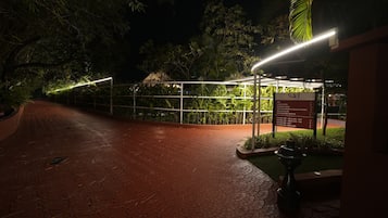 Um caminho pavimentado leva à entrada de um edifício, com uma placa de identificação, cercado por vegetação e área bem iluminada.