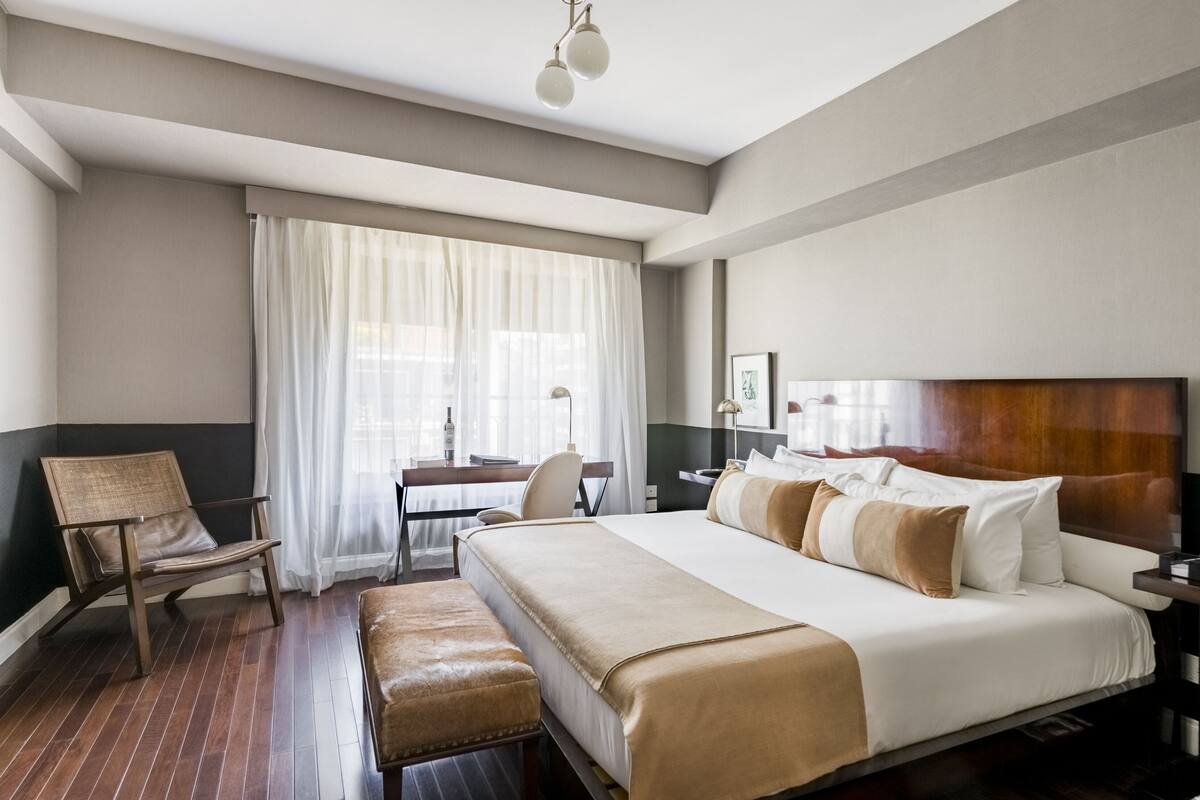 Quarto de hotel com uma cama grande, uma cadeira de madeira, uma escrivaninha e uma janela com cortinas leves.