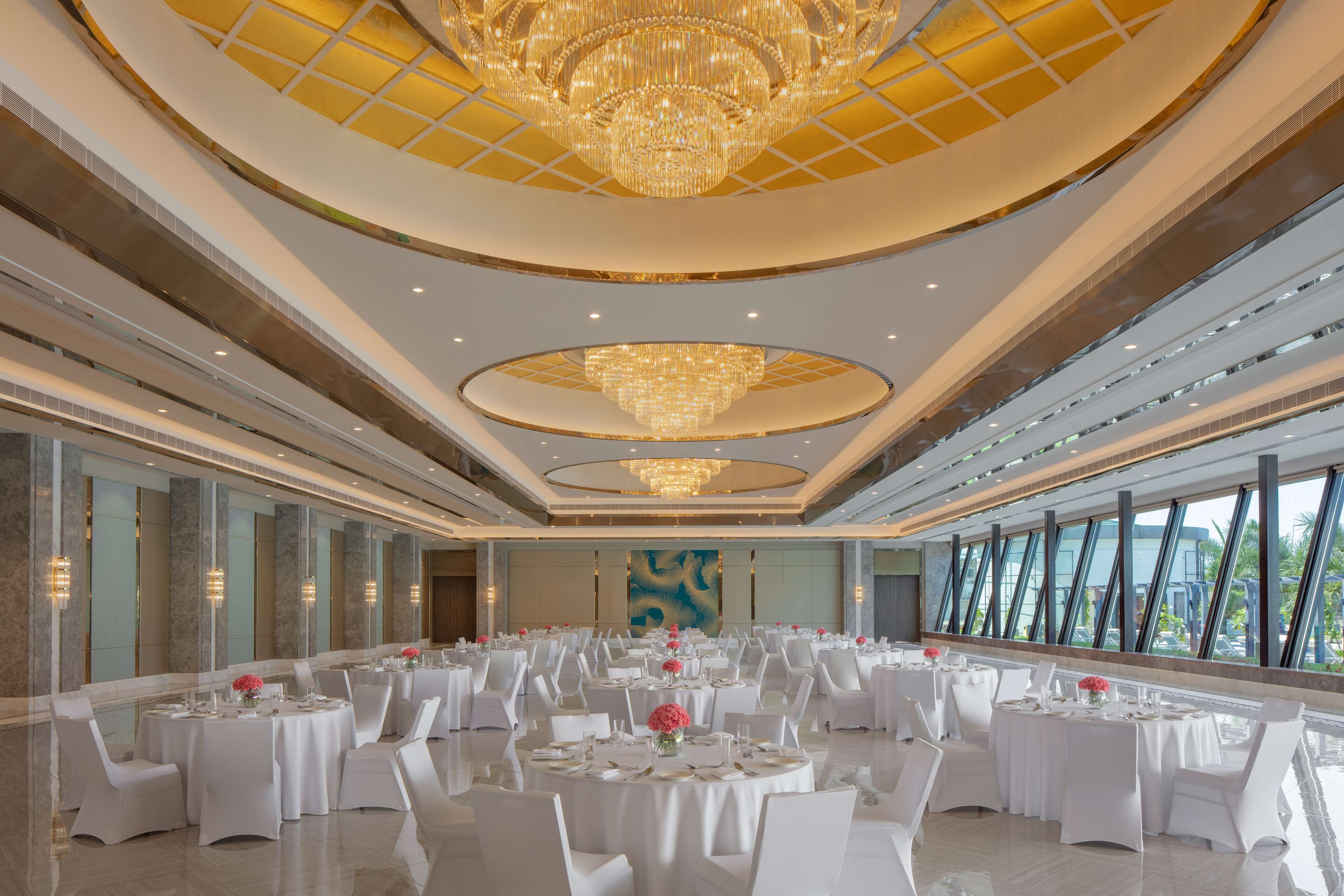 Um amplo salão de banquetes com várias mesas redondas preparadas para refeições, grandes janelas com vista e um imponente candelabro.