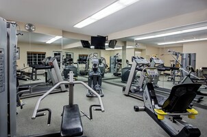 Academia com diversos equipamentos de exercícios, espelhos e piso coberto de carpete.