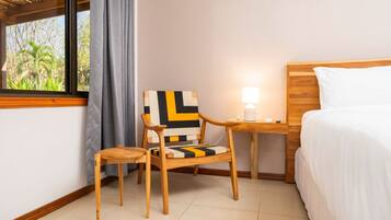 Quarto de hotel com cama, uma cadeira, uma mesinha com abajur e janela com vista para árvores.