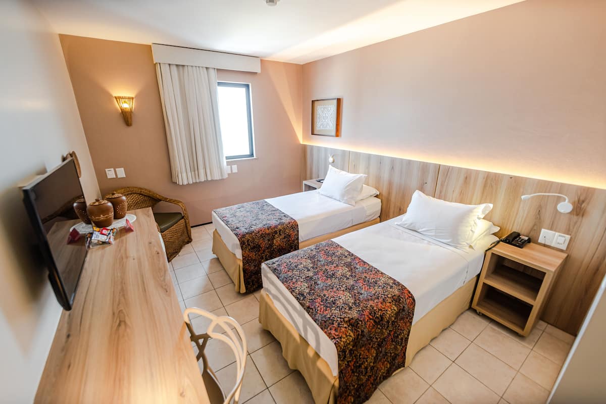 Quarto de hotel com duas camas, uma mesa de escritório, uma cadeira, uma televisão e uma mesa de cabeceira com abajur.