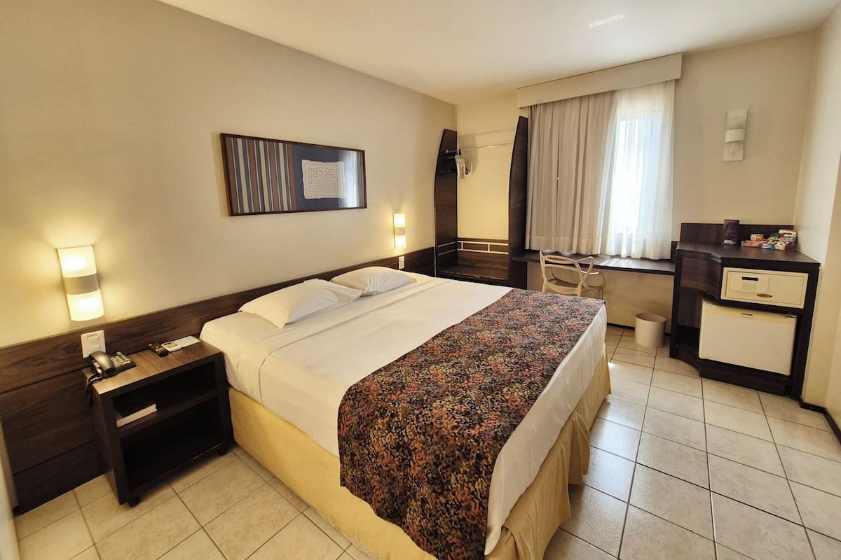 Quarto de hotel com cama, mesa de cabeceira, luminária, escrivaninha, cadeira e um pequeno frigorífico.