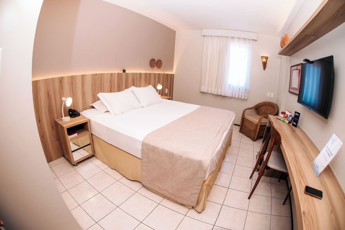 Quarto de hotel com cama, mesas de cabeceira, escrivaninha e cadeiras, televisão e janela com cortinas.