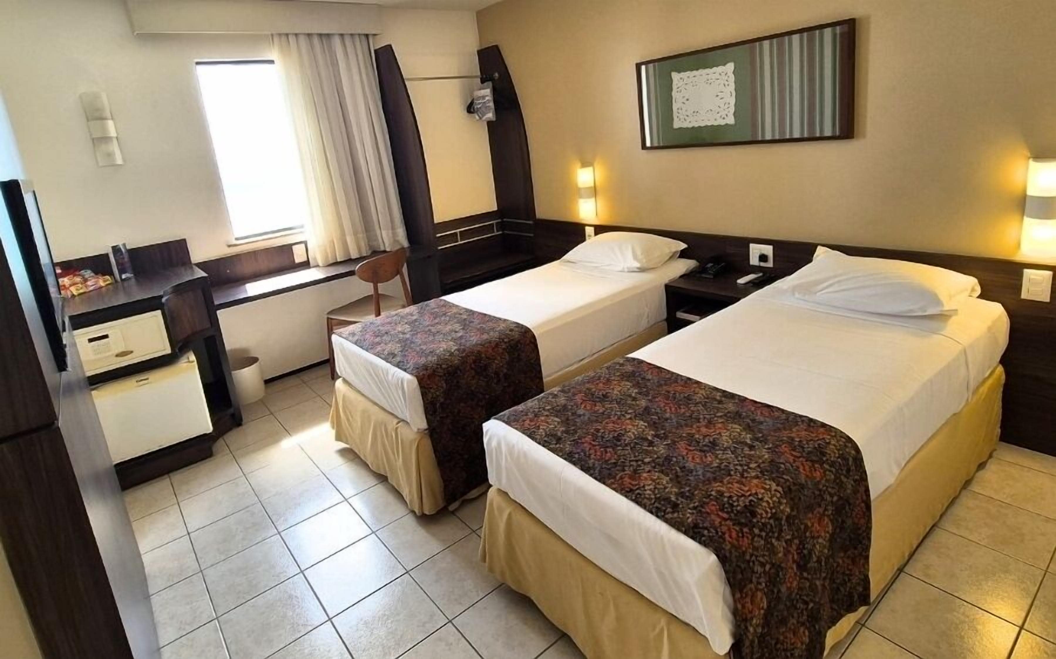 Quarto de hotel com duas camas, uma escrivaninha com cadeira, uma mesinha e uma janela com cortinas.