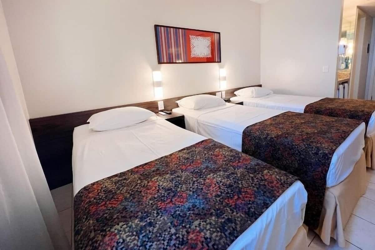 Quarto de hotel com três camas de solteiro, cada uma com uma colcha estampada, uma mesinha de cabeceira e uma luminária fixada na parede.