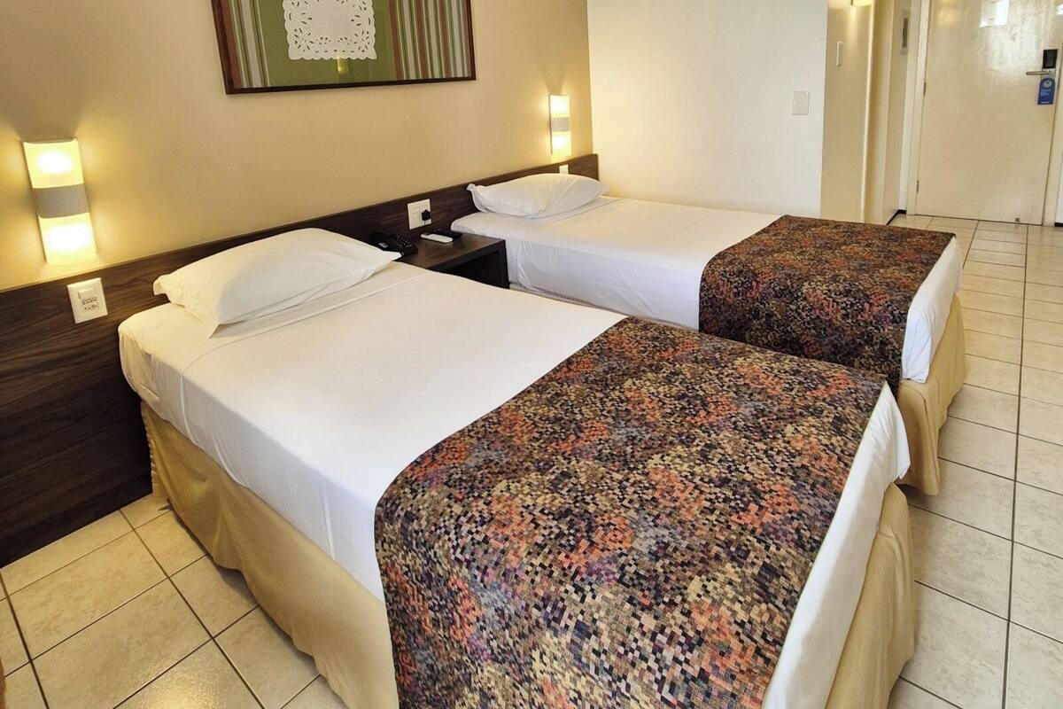 Quarto de hotel com duas camas, cada uma com lençóis brancos e um manta decorada. Há uma obra emoldurada acima das camas e uma luminária na parede à esquerda.