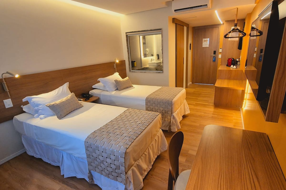 Quarto de hotel com duas camas, cabeceiras de madeira, um espelho, uma mesa de madeira e um banco.