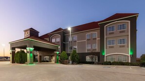 Um hotel com vários andares, área de estacionamento coberta, entrada visível e um jardim frontal ajardinado.