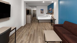 Quarto de hotel com cama, um sofá marrom, televisão, escrivaninha e uma mesa pequena.