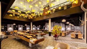Uma ampla área de refeições com várias mesas e cadeiras, um buffet com diversos alimentos e uma grande planta em vaso com flores amarelas.