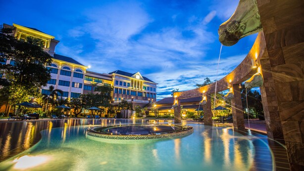 Uma área de piscina de hotel com uma fonte, cercada por edifícios e palmeiras.