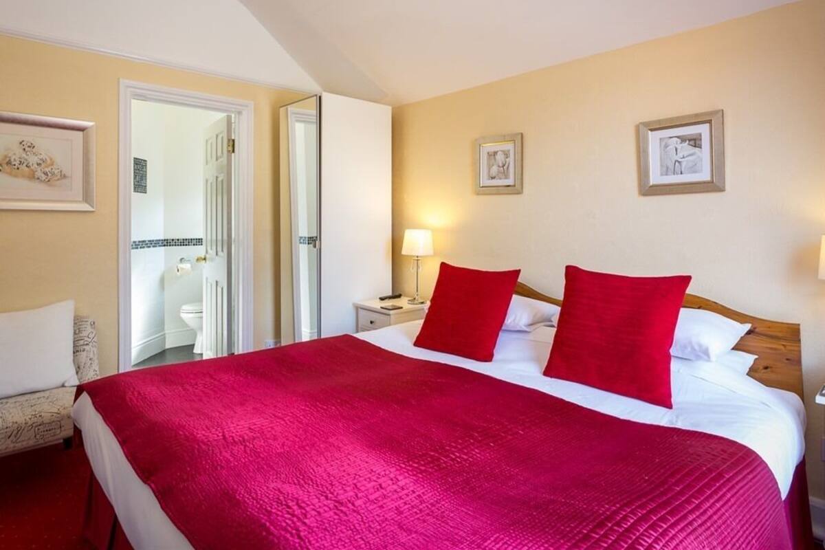 Quarto de hotel com uma cama grande, duas almofadas vermelhas, cabeceira de madeira, mesa de cabeceira com abajur e quadros nas paredes.