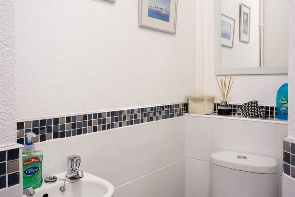 Casa de banho com lavatório branco, sanita e espelho sobre o lavatório. Existem quadros nas paredes e um painel de azulejos azul e branco.