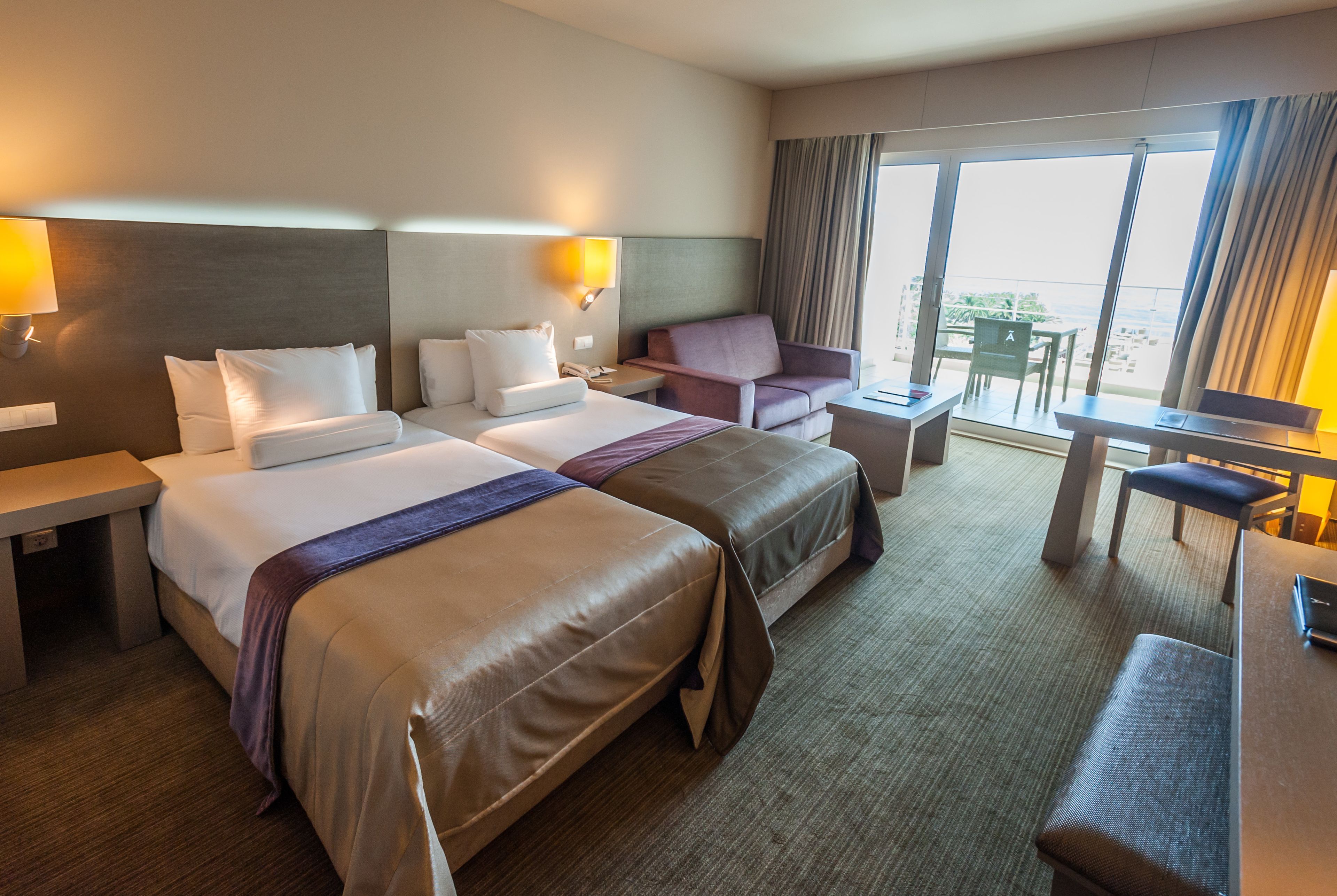 Quarto de hotel com duas camas, uma escrivaninha, uma cadeira e varanda com vista.