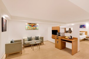 Quarto de hotel moderno com sofá, mesa de centro, escrivaninha com televisão de tela plana, micro-ondas e cama com cabeceira.